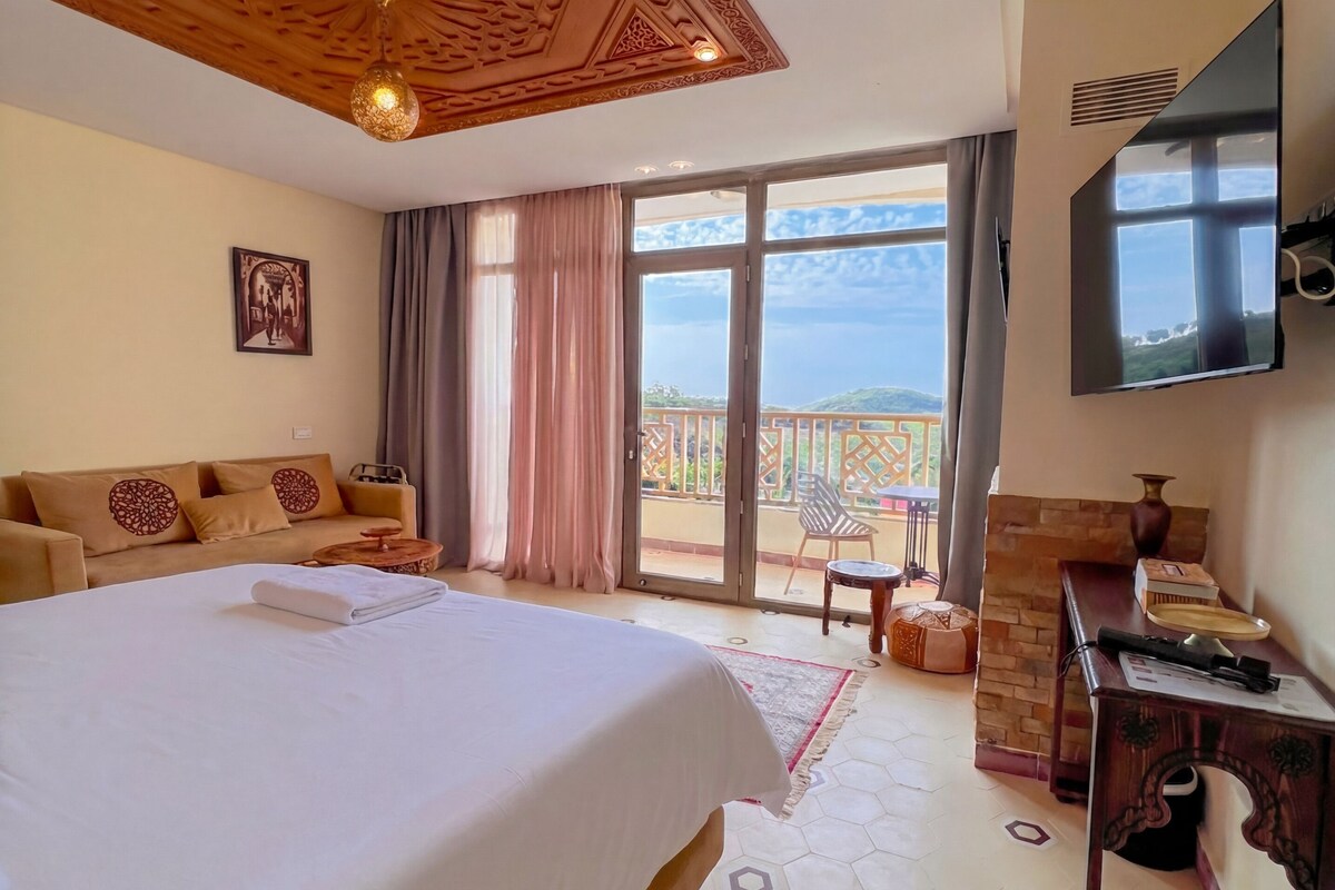 Quarto de hotel com uma cama grande, um sofá, uma TV e uma varanda com vista.