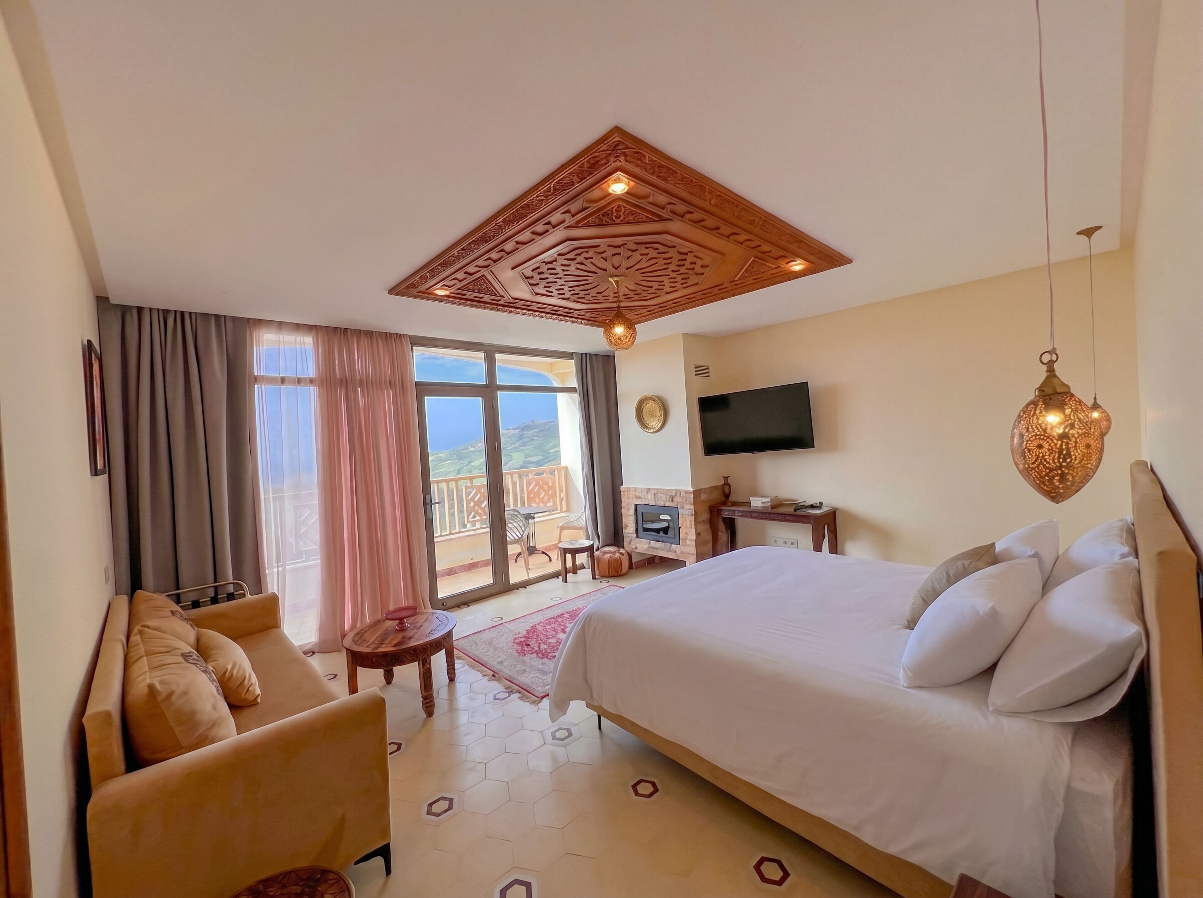 Quarto de hotel com cama, sofá, televisão, lareira e varanda com vista.