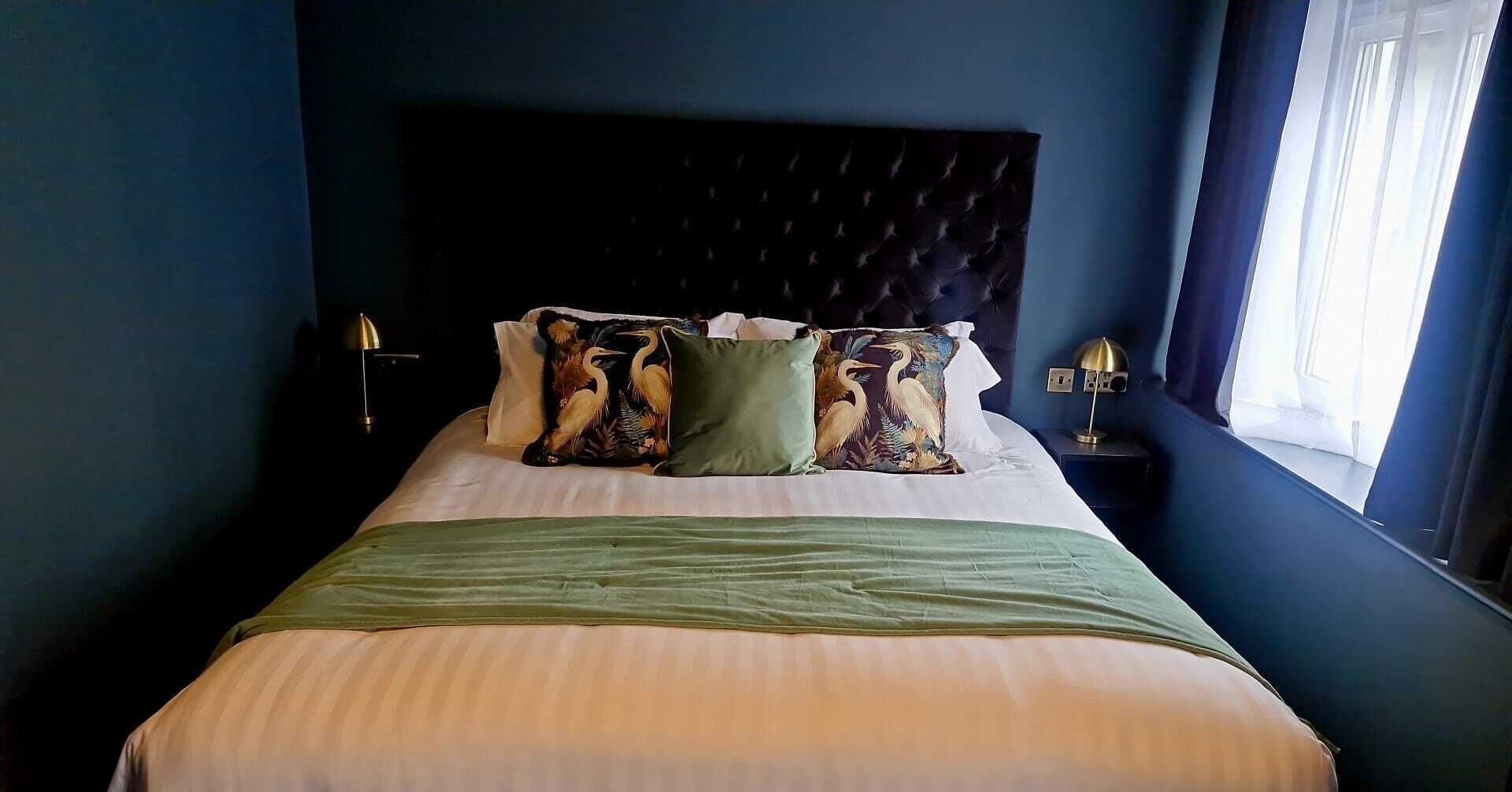 Cama bem arrumada com colcha verde e almofadas decorativas, cabeceira escura e mesa de cabeceira com abajur.