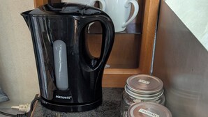 Uma área de cozinha com uma cafeteira Muenett preta, quatro potes de vidro identificados com diferentes conteúdos e uma prateleira com copos brancos.