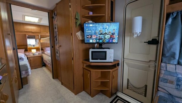 Interior de uma RV compacta, com televisão, armários de madeira e uma pequena cozinha.