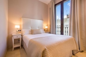 Quarto de hotel com uma cama grande, duas mesas de cabeceira, uma janela com cortinas e vista para edifícios.