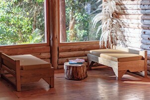 Interior de uma cabana de madeira com um banco, uma pequena mesa com livros e uma janela com vista para o verde.