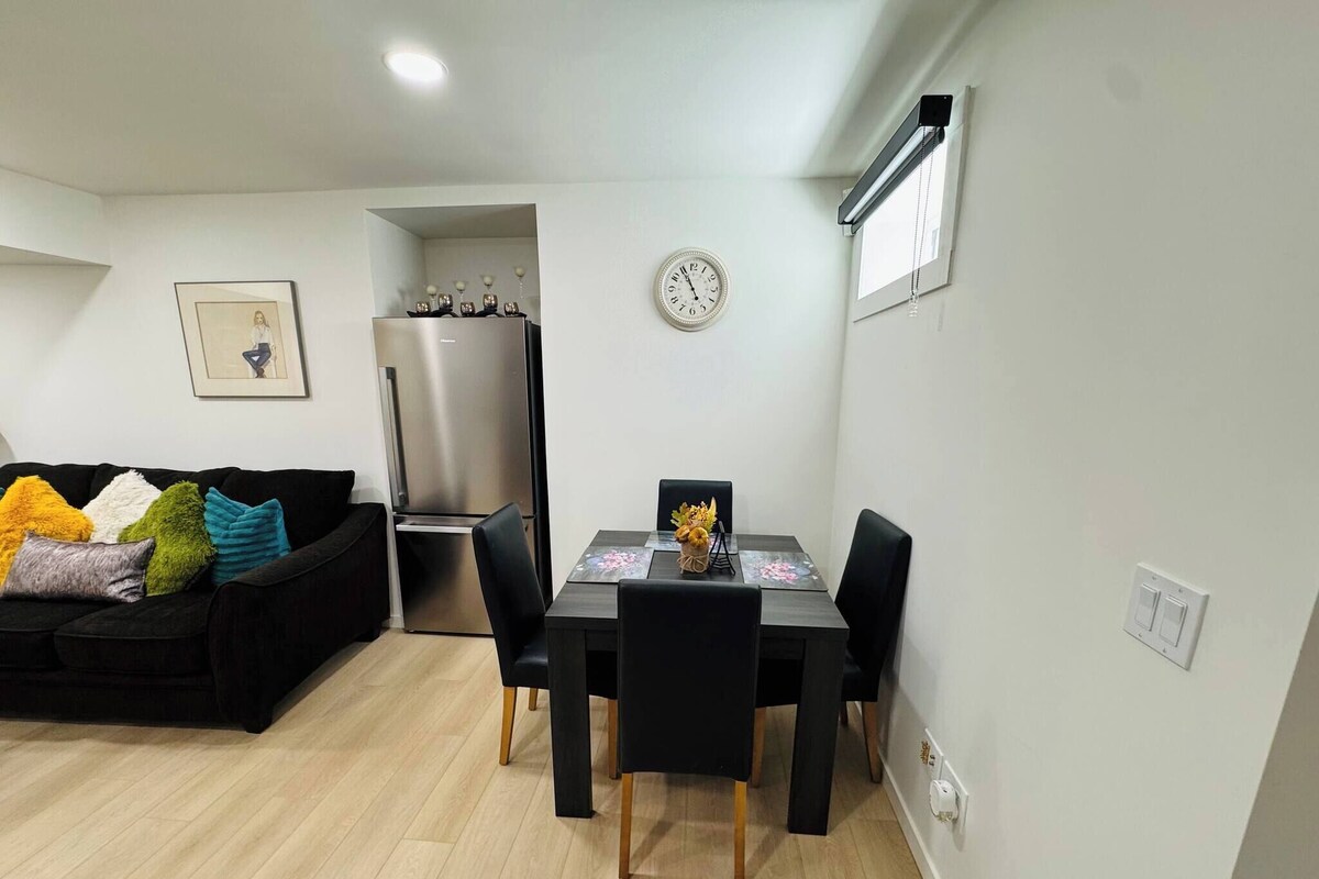 Uma sala de estar moderna com um sofá preto, uma mesa de jantar com cadeiras, uma geladeira e um relógio de parede.