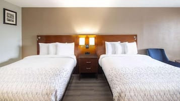 Quarto de hotel com duas camas, uma mesinha de cabeceira, uma luminária e uma cadeira.