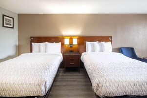 Quarto de hotel com duas camas, uma mesinha de cabeceira, uma luminária e uma cadeira.