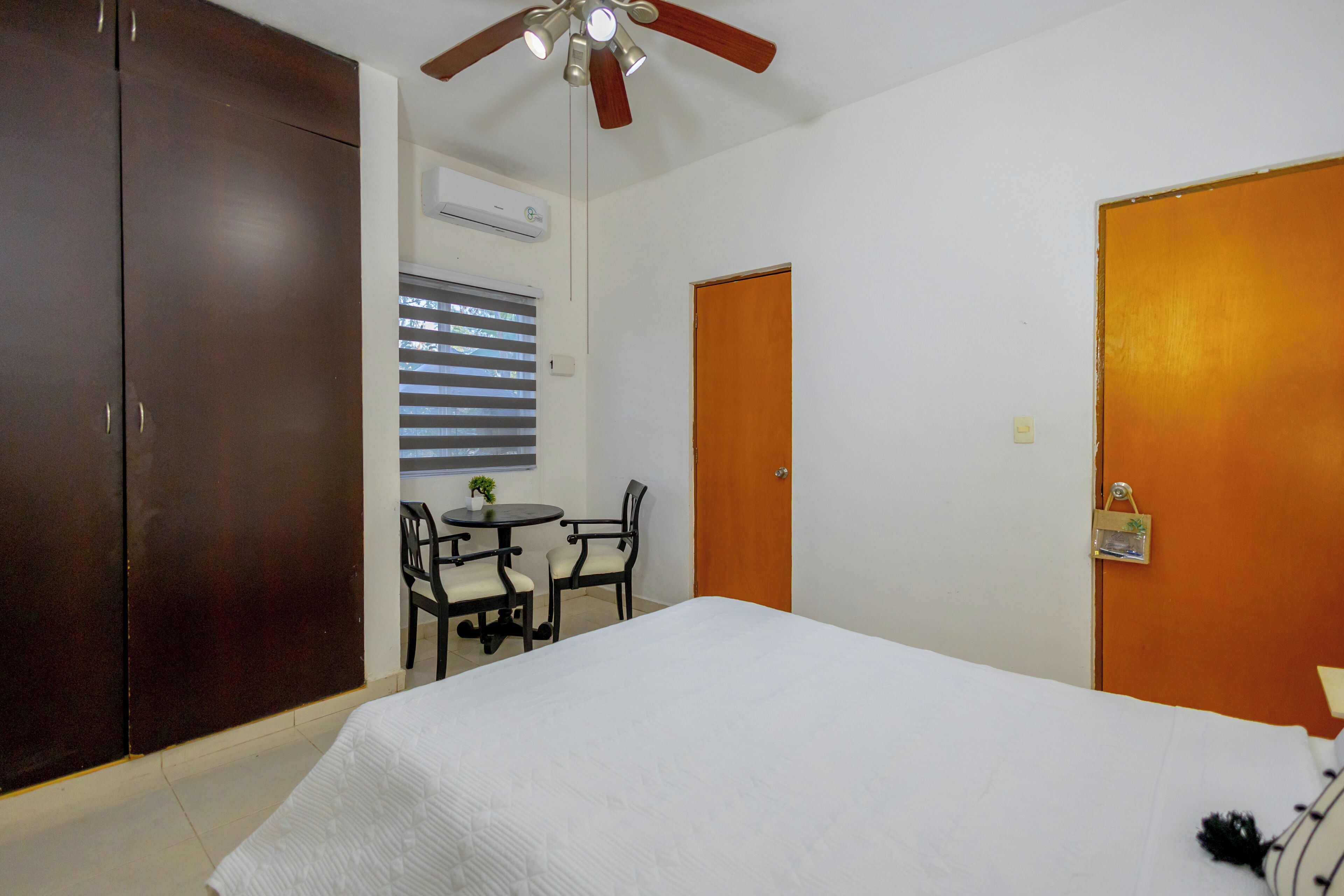 Quarto com cama, ventoinha no teto, janela com persianas, mesa pequena com cadeiras e guarda-roupa.