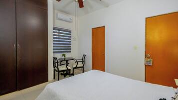 Quarto com cama, ventoinha no teto, janela com persianas, mesa pequena com cadeiras e guarda-roupa.