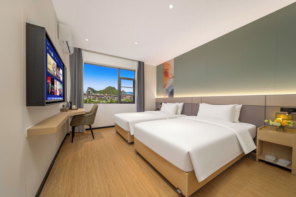 Quarto de hotel com duas camas, uma escrivaninha, uma televisão e janela com vista.
