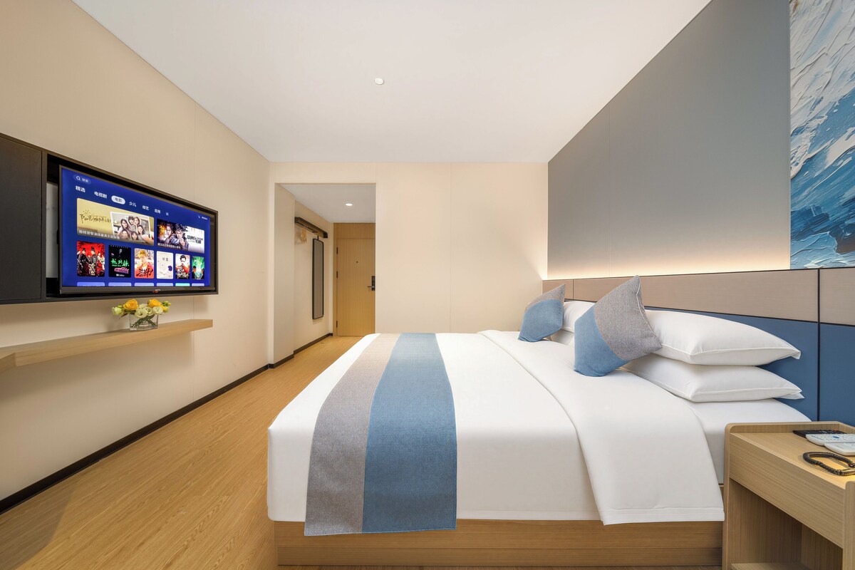 Quarto de hotel com cama, televisão presa na parede, piso de madeira e um quadro na parede.