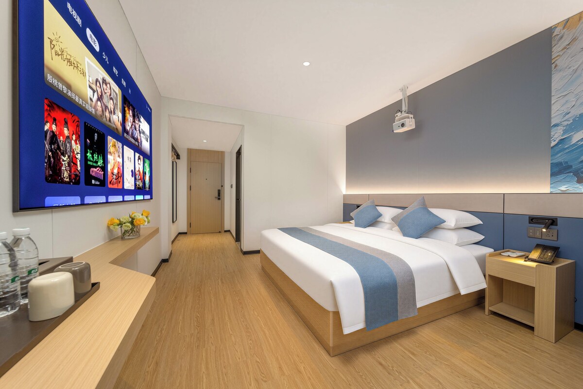 Quarto de hotel com cama grande, mesa de cabeceira com telefone, escrivaninha com computador e garrafas de água, TV na parede exibindo pôsteres de filmes, e corredor que leva a outro quarto.