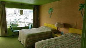 Quarto de hotel com duas camas, uma mesa pequena, uma cadeira e janela com vista para palmeiras e edifícios.