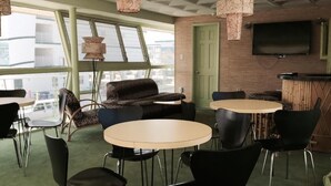 Um quarto com mesas e cadeiras redondas, um sofá, uma televisão e grandes janelas com vista para prédios.