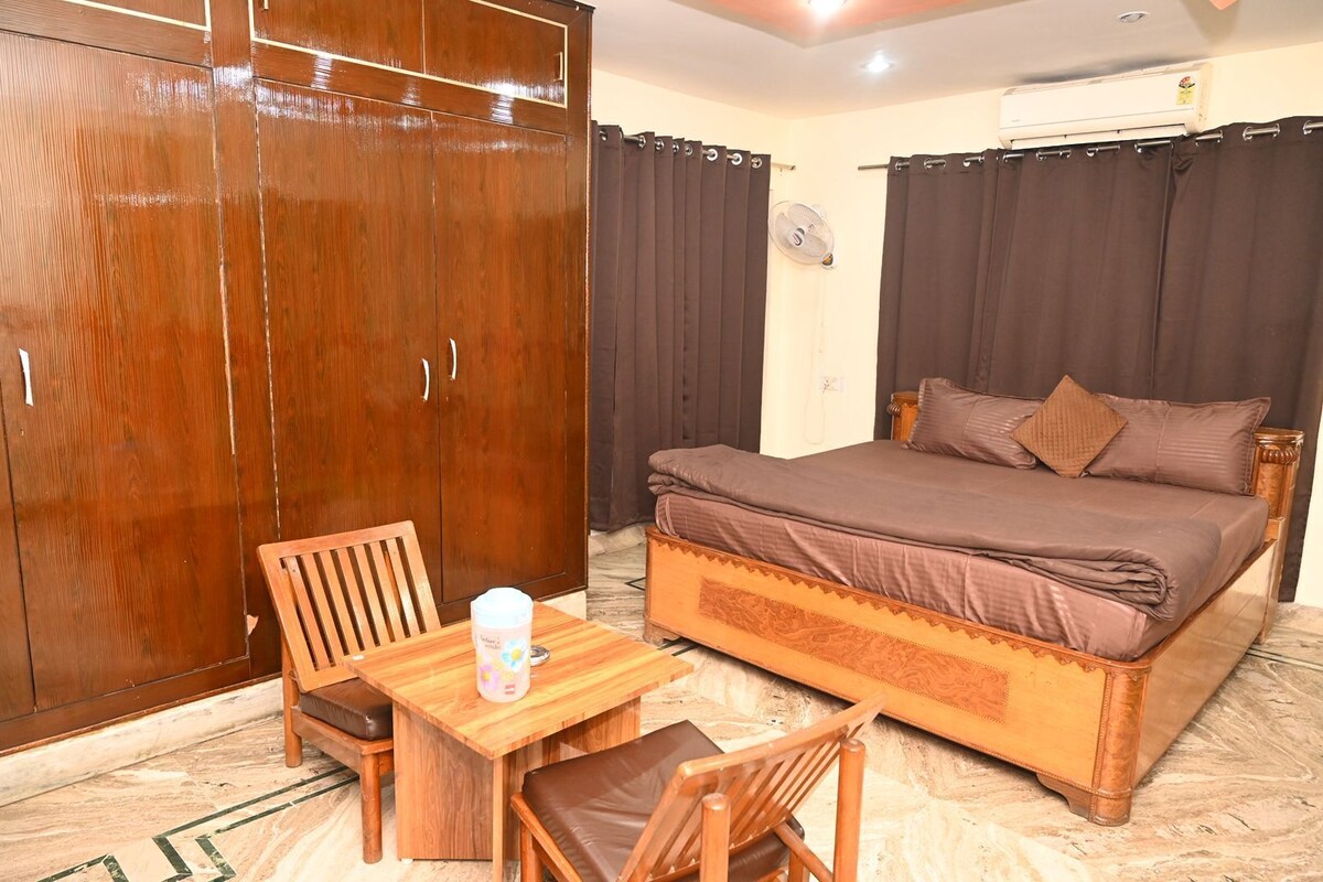 Quarto com cama, guarda-roupa de madeira, mesa e cadeiras, e um ventilador.