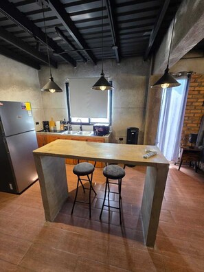 Cozinha com ilha central, dois banquetes, iluminação pendente, geladeira e parede de tijolos.