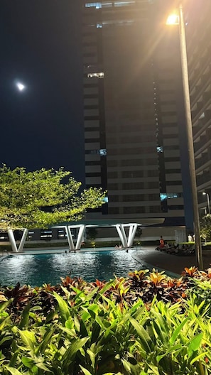 Vista noturna de um edifício alto com área de piscina em primeiro plano, apresentando uma ponte e vegetação exuberante.