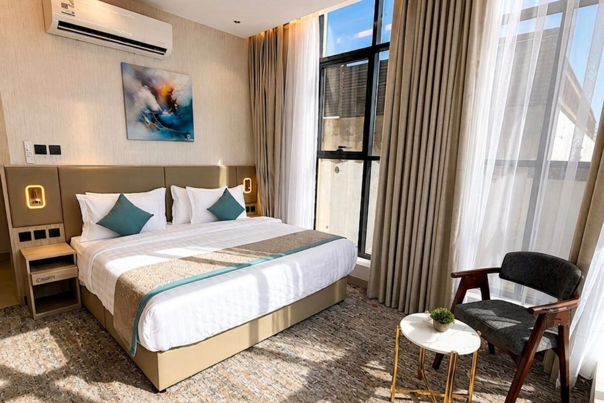 Quarto de hotel moderno com uma cama grande, uma mesinha, uma cadeira e uma janela com cortinas.