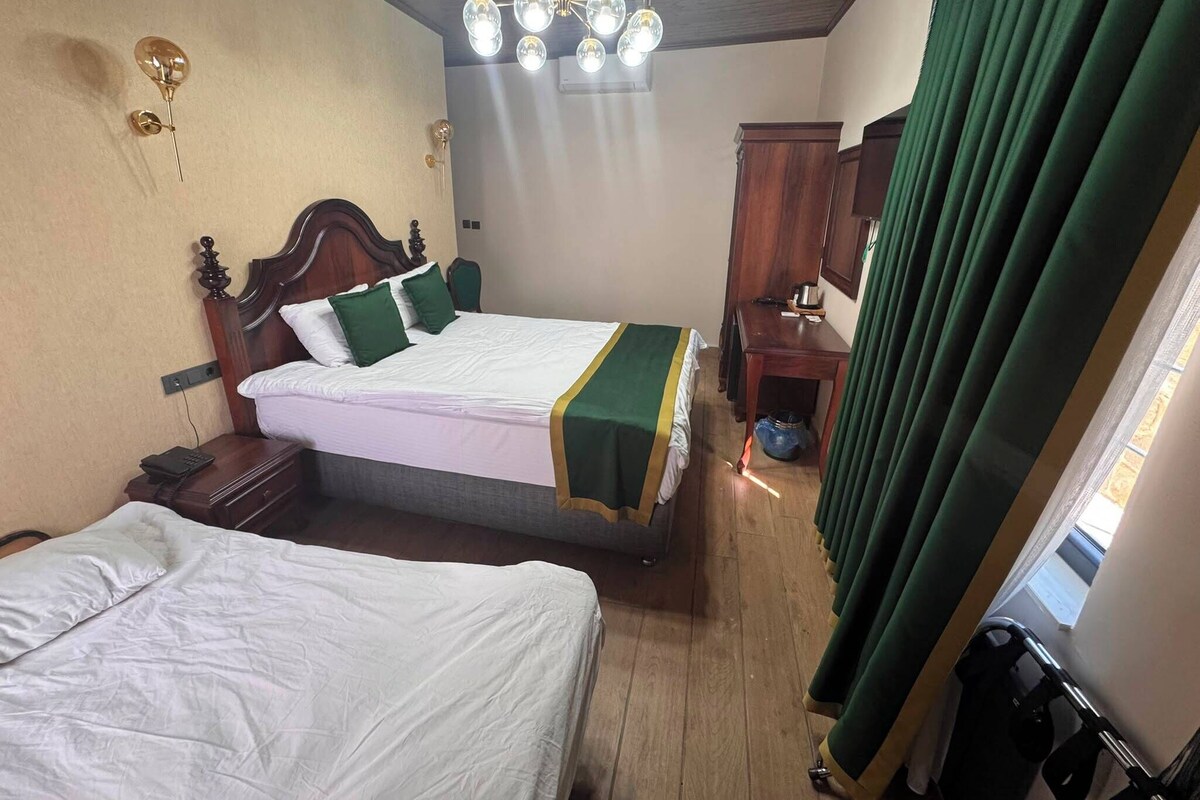 Quarto de hotel com cama de casal, mesa de cabeceira, escrivaninha, cadeira, guarda-roupa e cortina verde.