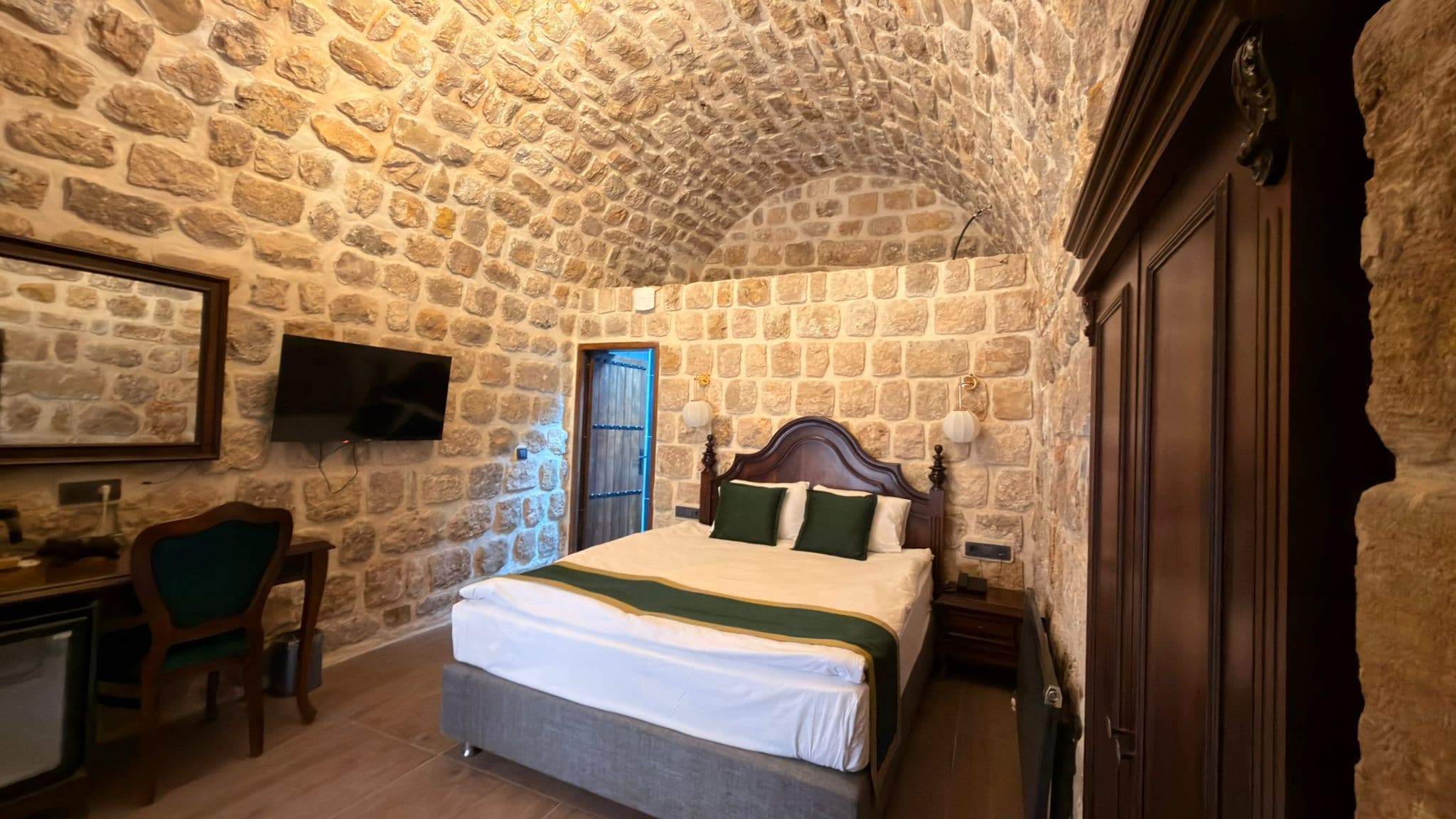 Quarto com parede de pedra, cama com roupa de cama verde e branca, escrivaninha com cadeira, televisão e guarda-roupa de madeira.