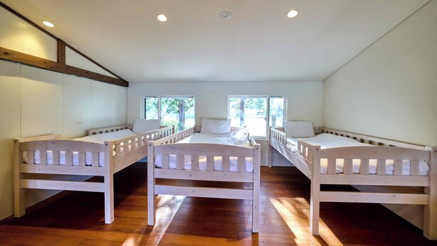 Quarto com três camas de madeira dispostas em linha, teto inclinado e grandes janelas que permitem entrada de luz natural.