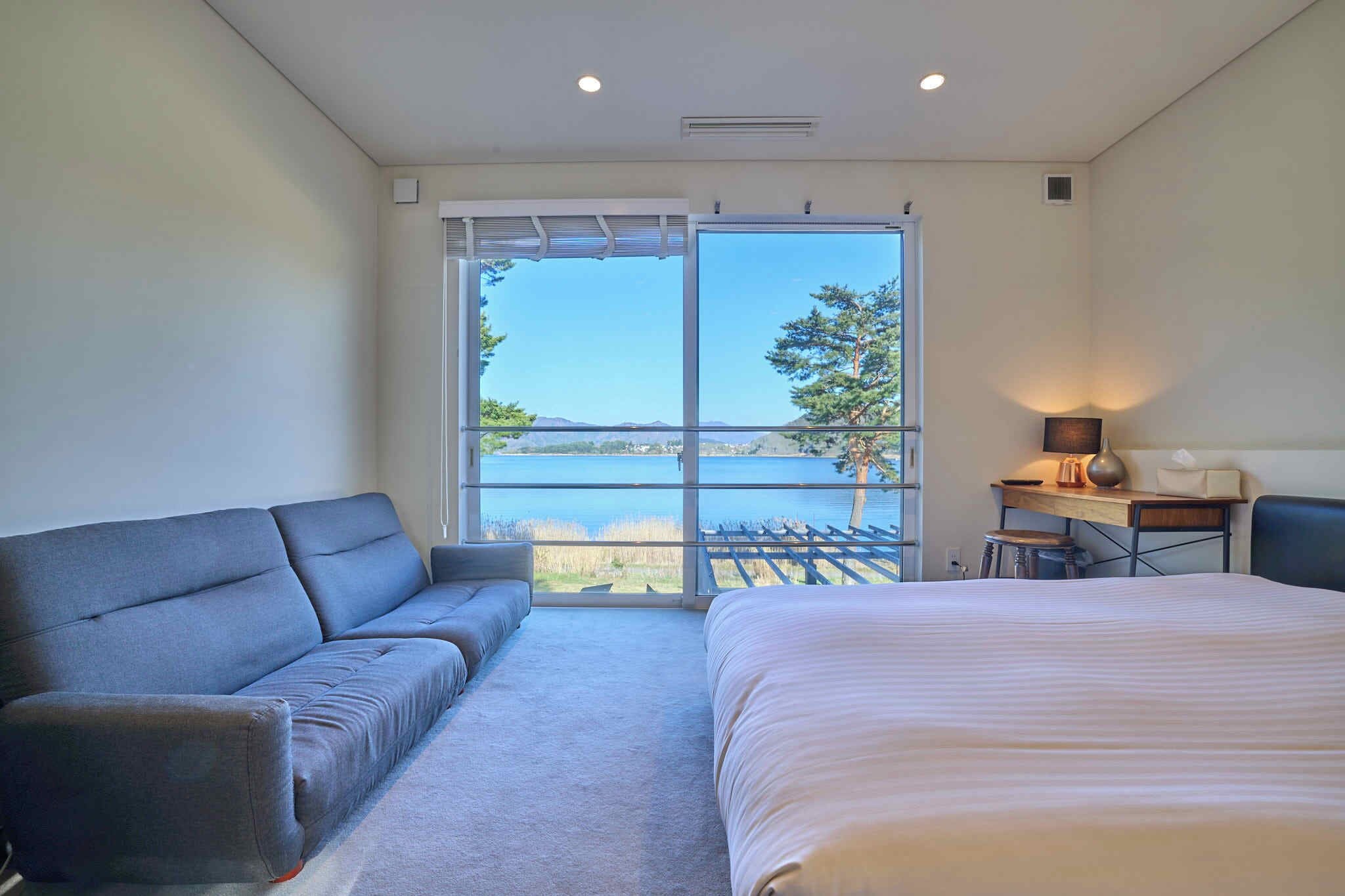 Quarto com cama, sofá, escrivaninha com abajur e vista para um lago através de grandes janelas.