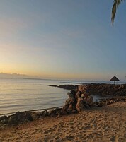 Uma praia com formação rochosa, um guarda-sol de palha e um mar calmo.