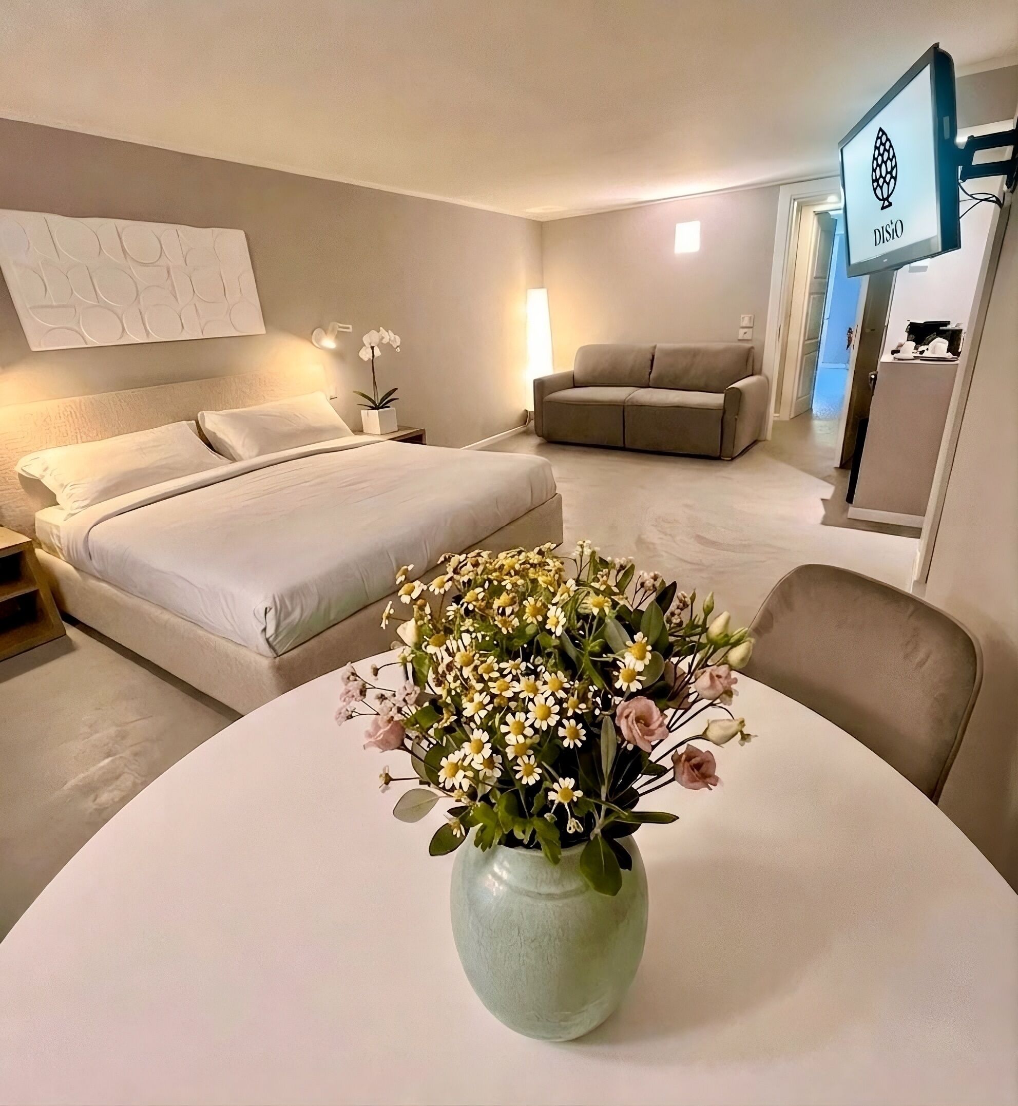 Quarto de hotel com uma cama, um sofá, uma televisão e um vaso com flores sobre uma mesa.