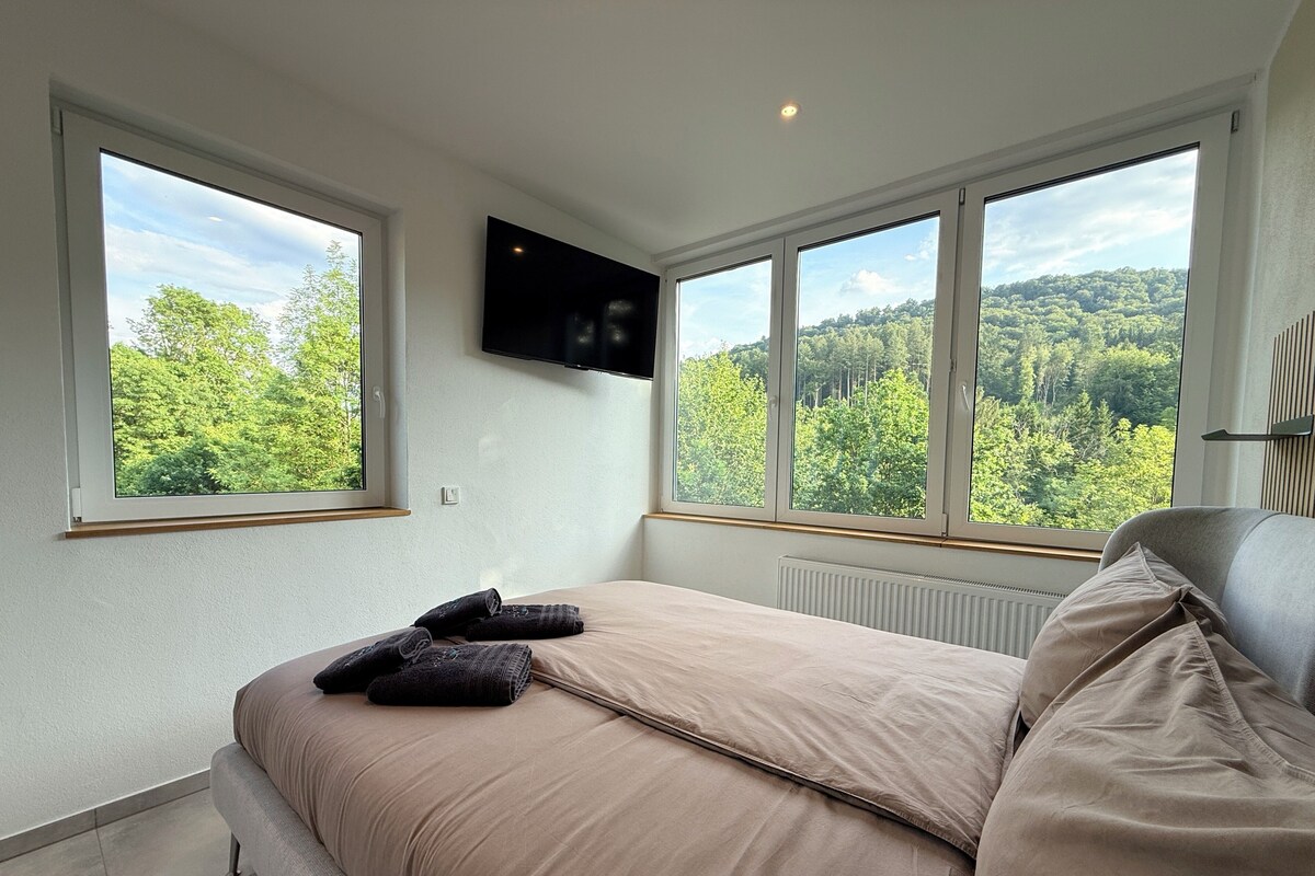 Quarto com cama, TV fixada na parede e amplas janelas com vista para uma área arborizada.