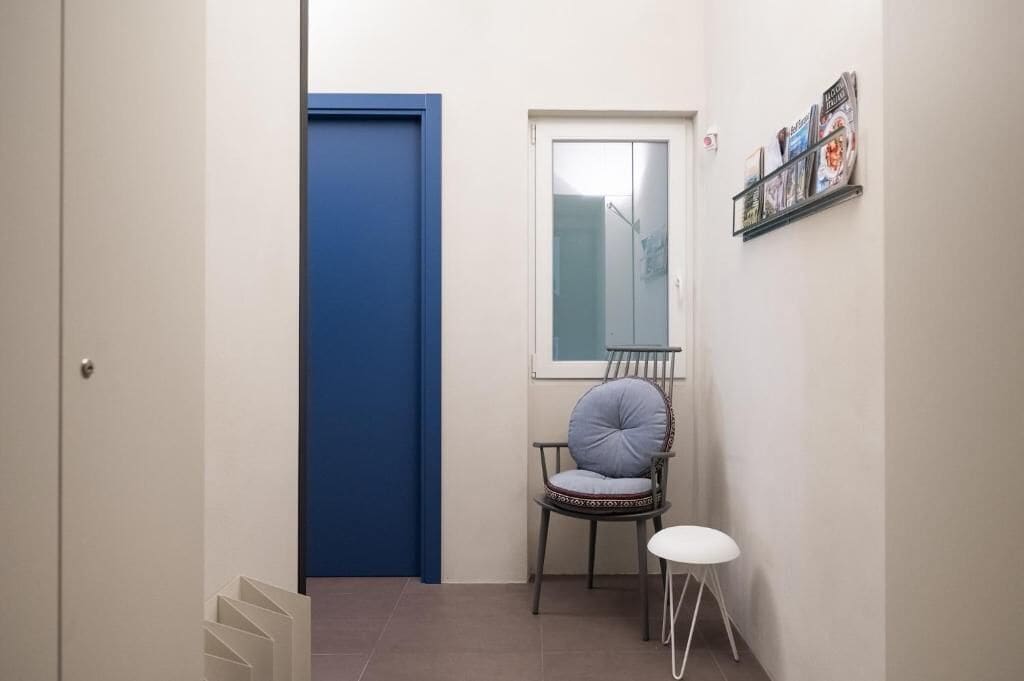 Um corredor estreito com uma porta azul, uma cadeira com almofada, um banquinho branco pequeno e uma janela com vista.