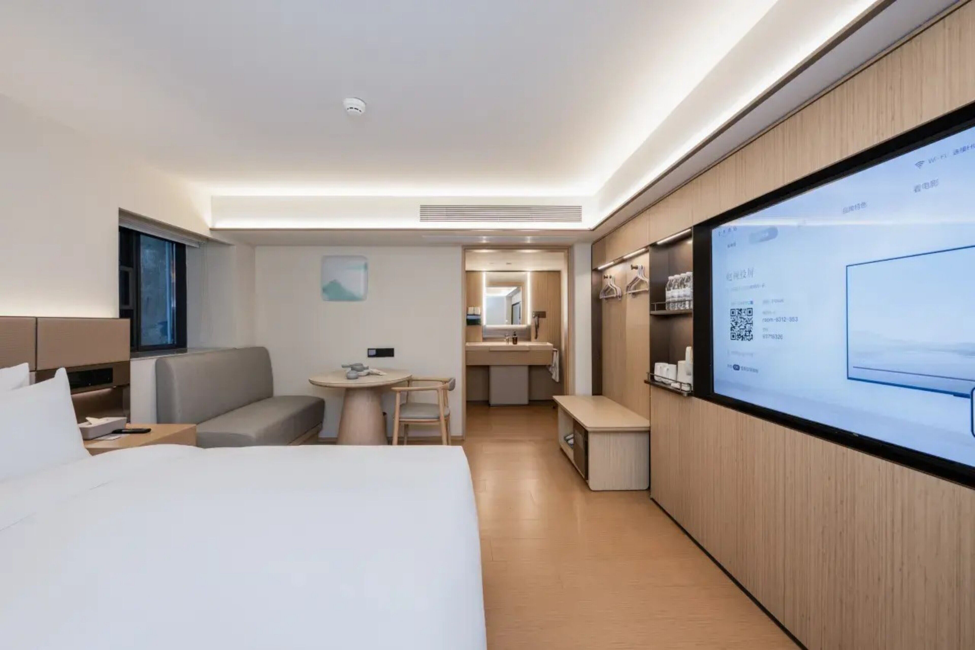 Quarto de hotel moderno com cama, área de estar, escrivaninha, espelho e TV exibindo informações.