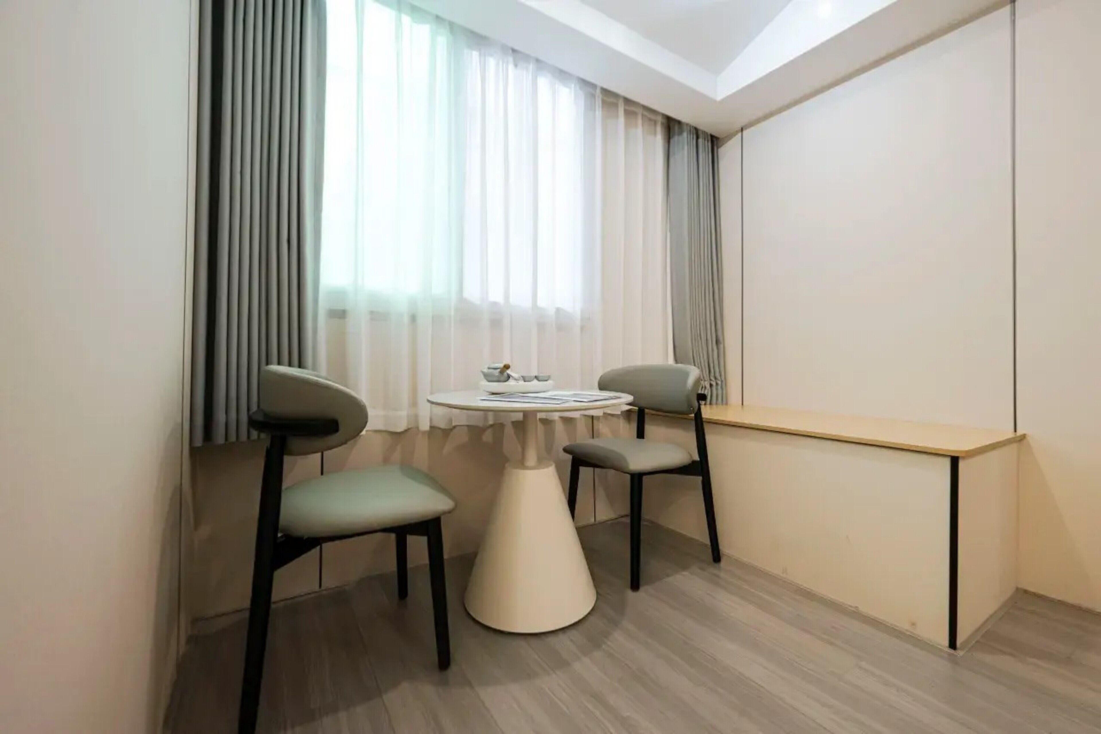 Um quarto pequeno e moderno, com uma mesa redonda, duas cadeiras, uma escrivaninha e uma janela com cortinas leves.