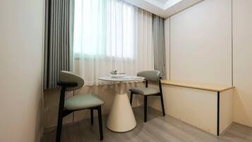 Um quarto pequeno e moderno, com uma mesa redonda, duas cadeiras, uma escrivaninha e uma janela com cortinas leves.