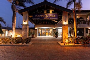 Entrada do Hotel Dorado Eagle Beach com galeria coberta, palmeiras e caminho de tijolos.