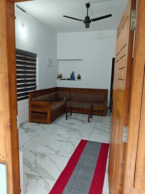 Sala de estar com sofá seccional, ventilador de teto, tapete cinza e vermelho, e prateleira fixada na parede com objetos decorativos.