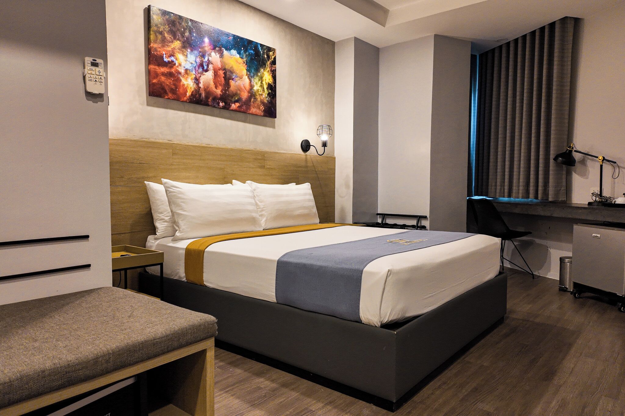 Quarto de hotel com cama, mesas de cabeceira, escrivaninha, cadeira, luminária de mesa, lixeira e uma grande pintura abstrata na parede.