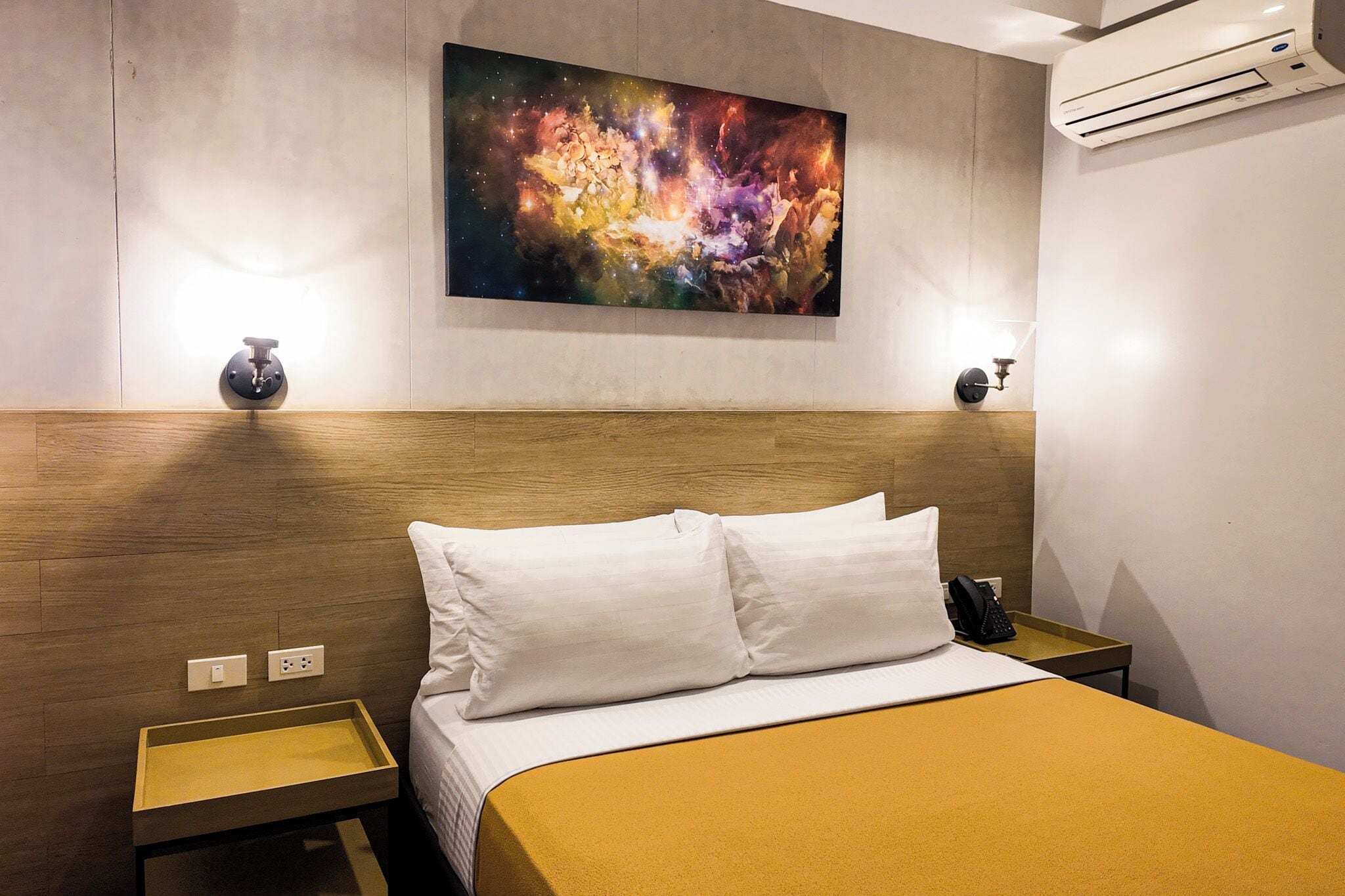 Quarto de hotel com cama, mesas de cabeceira, ar condicionado fixado na parede e um quadro abstrato grande.