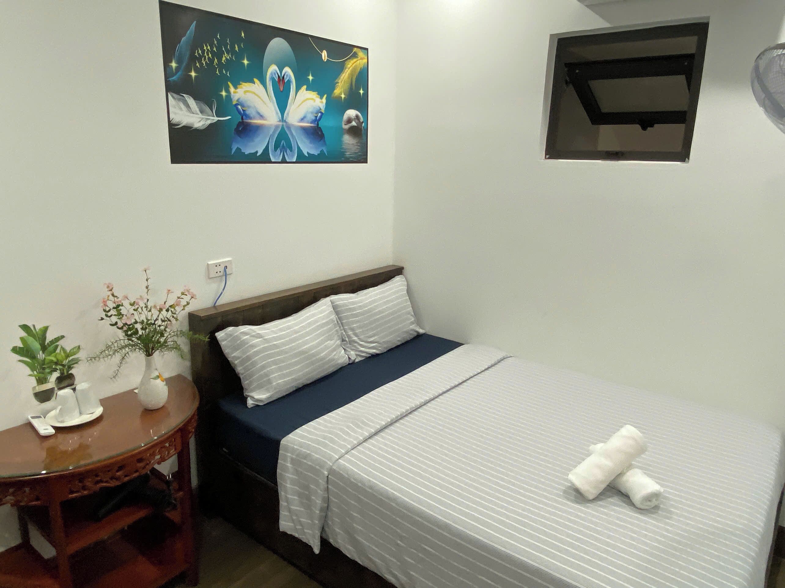 Quarto com cama, mesa de cabeceira com vaso e planta, e um quadro na parede.
