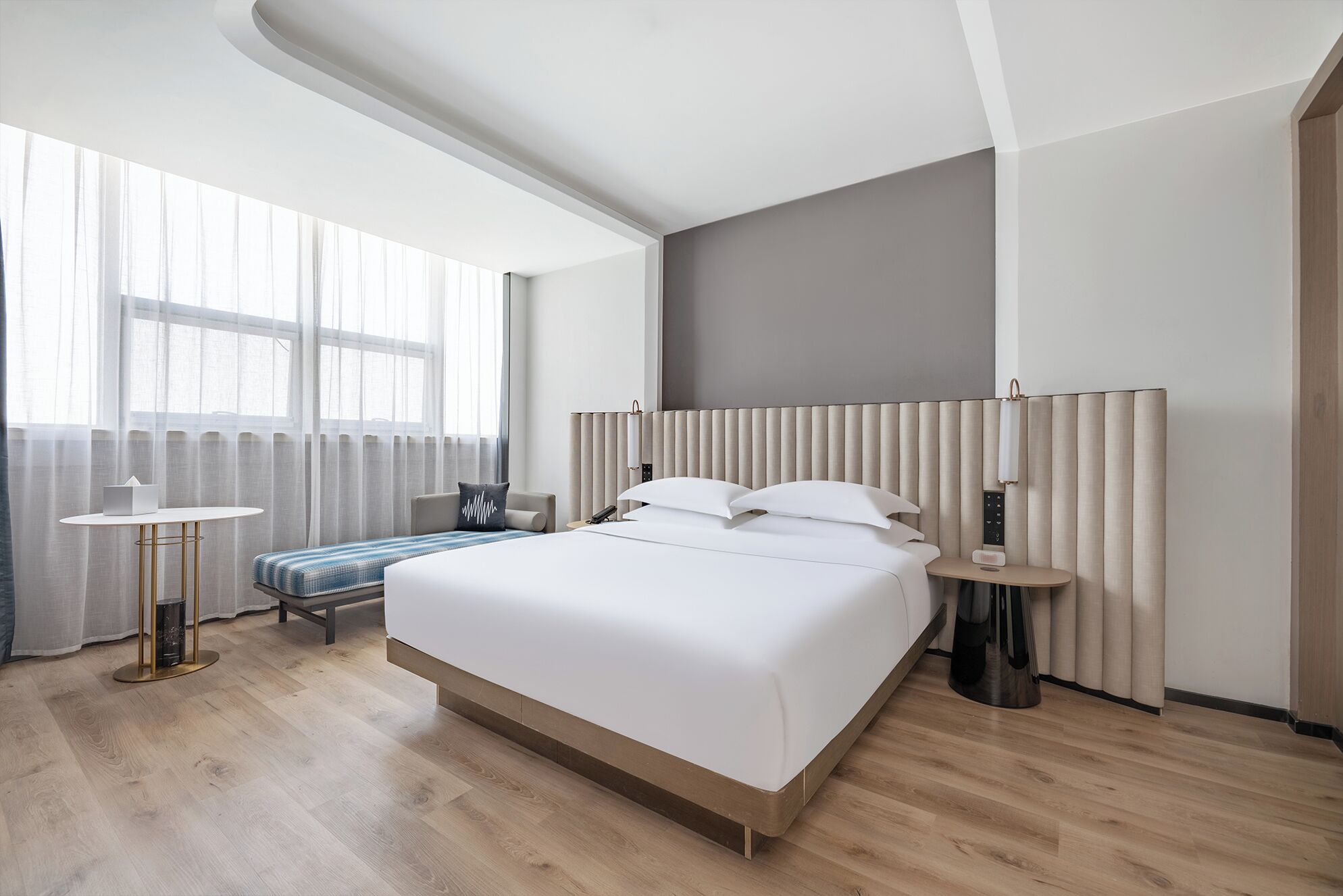 Quarto de hotel moderno com uma cama grande, mesas de cabeceira, uma pequena mesa redonda e uma janela com cortinas leves.