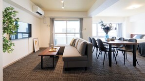 Quarto de hotel moderno com sofá, mesa de jantar, cadeiras, televisão e uma janela grande.
