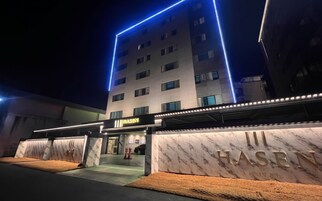 Entrada de hotel com uma placa 'HASEN', uma galeria coberta e um edifício com fachada iluminada em azul.