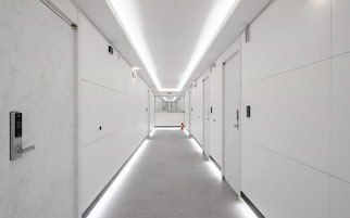 Corredor de hotel comprido e estreito, com paredes brancas e portas de ambos os lados, que termina numa parede espelhada.