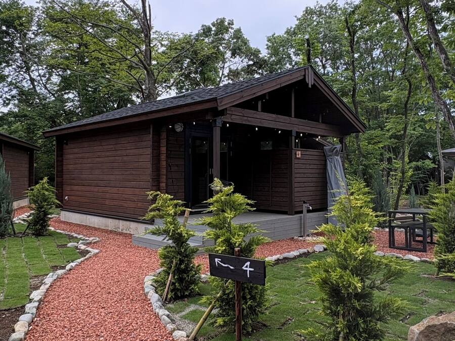 Uma cabana de madeira com um caminho de cascalho que a leva, rodeada por árvores e um pequeno jardim.