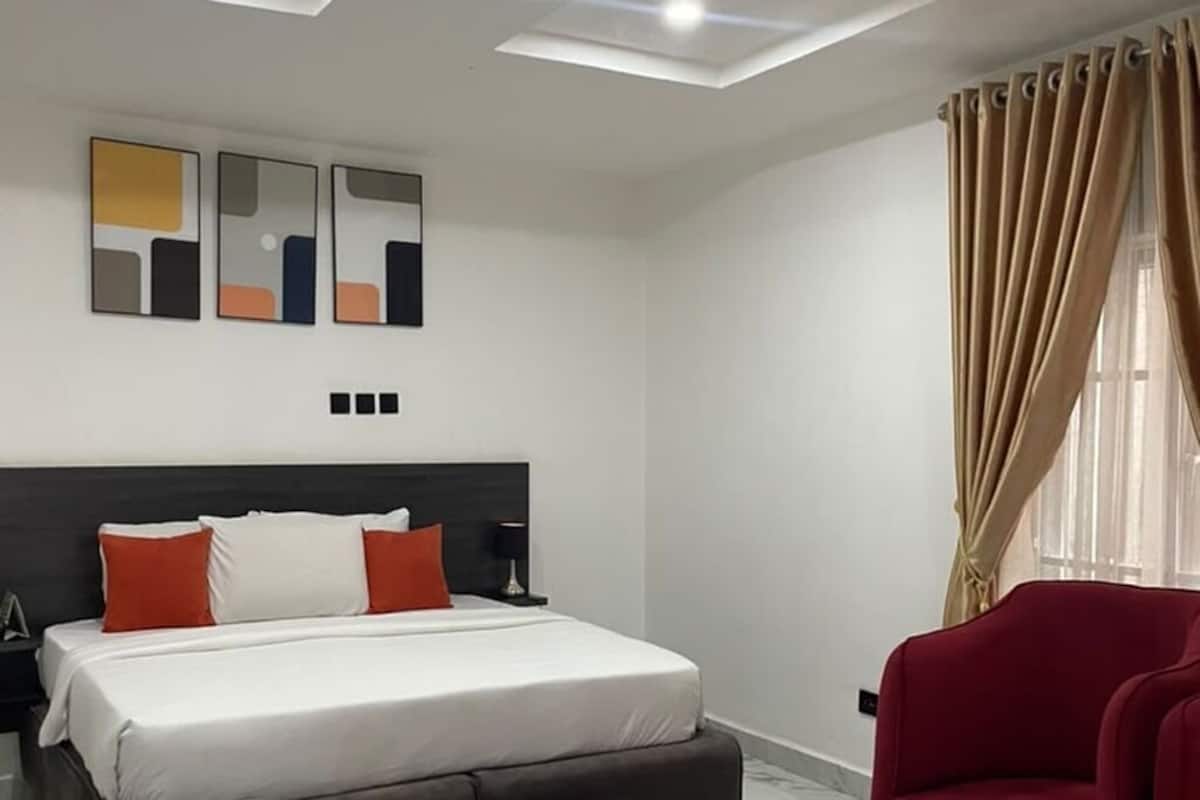 Quarto de hotel moderno com uma cama, uma poltrona vermelha, arte abstrata na parede e uma janela com cortinas.
