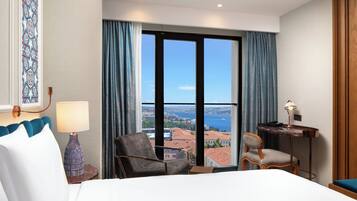 Quarto de hotel com uma cama grande, duas cadeiras, uma escrivaninha com abajur e vista para a cidade através de uma janela ampla.