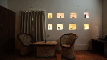 Quarto com duas cadeiras de vime, uma mesa pequena, cortina e luminárias fixadas na parede, além de diversos objetos decorativos.