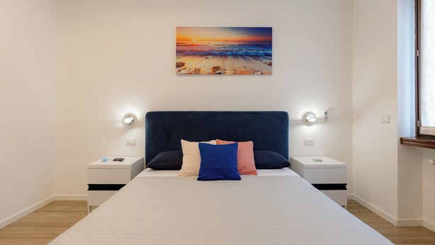 Quarto com uma cama, uma pintura de um pôr do sol, mesas de cabeceira e um criado-mudo.