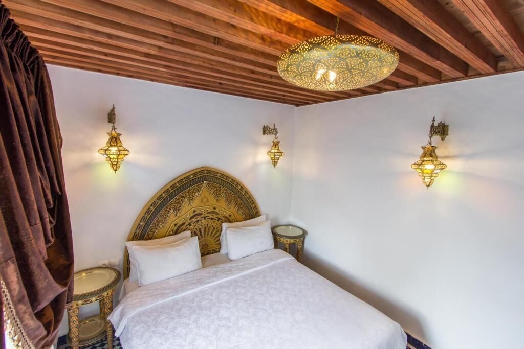 Uma cama com cabeceira decorativa, duas luminárias na parede e um lustre.