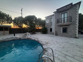 Piscina com escada de metal, edifício de pedra com varanda e pôr do sol ao fundo.