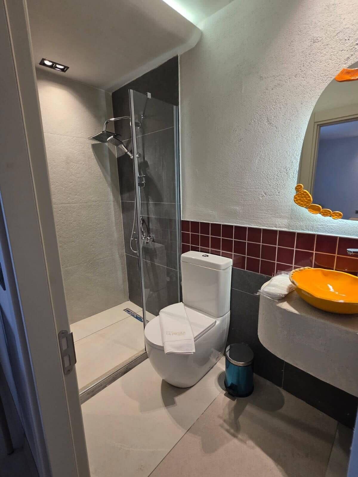 Uma casa de banho com cabine de duche em vidro, sanita branca, lavatório com cuba amarela e espelho com moldura decorativa.