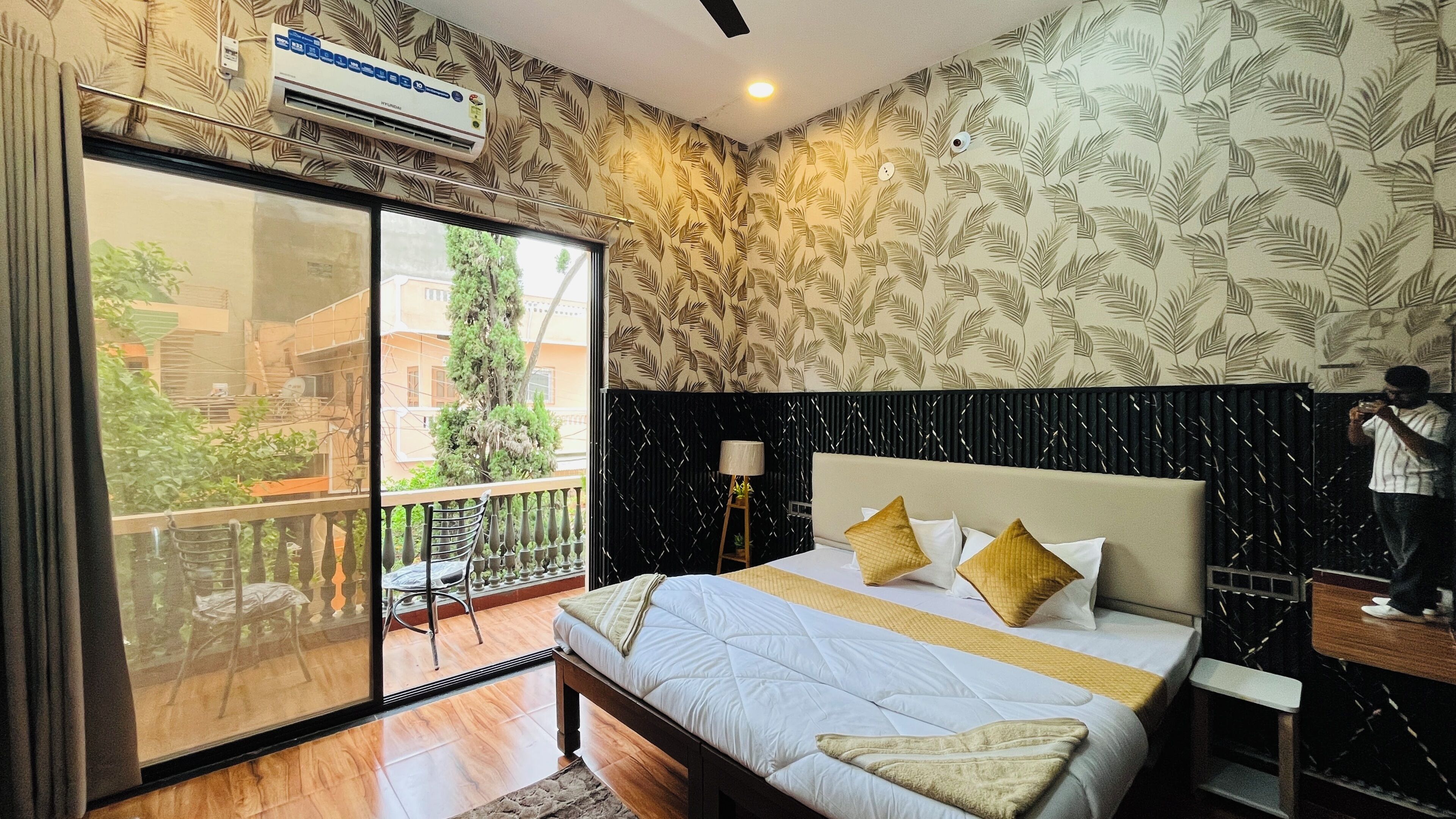 Quarto de hotel com cama, mesa de cabeceira, luminária e uma janela grande com vista para edifícios e vegetação.